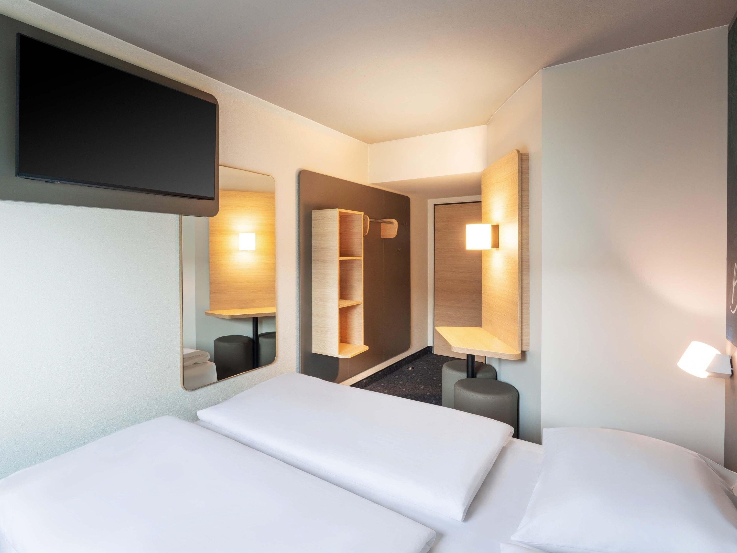 Quarto de hotel com cama, televisão fixada na parede e um espelho refletindo o ambiente.
