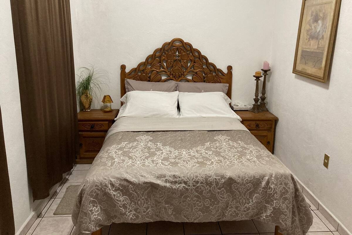 Cama bem arrumada com colcha estampada, cabeceira de madeira e mesas de cabeceira com abajures e planta.