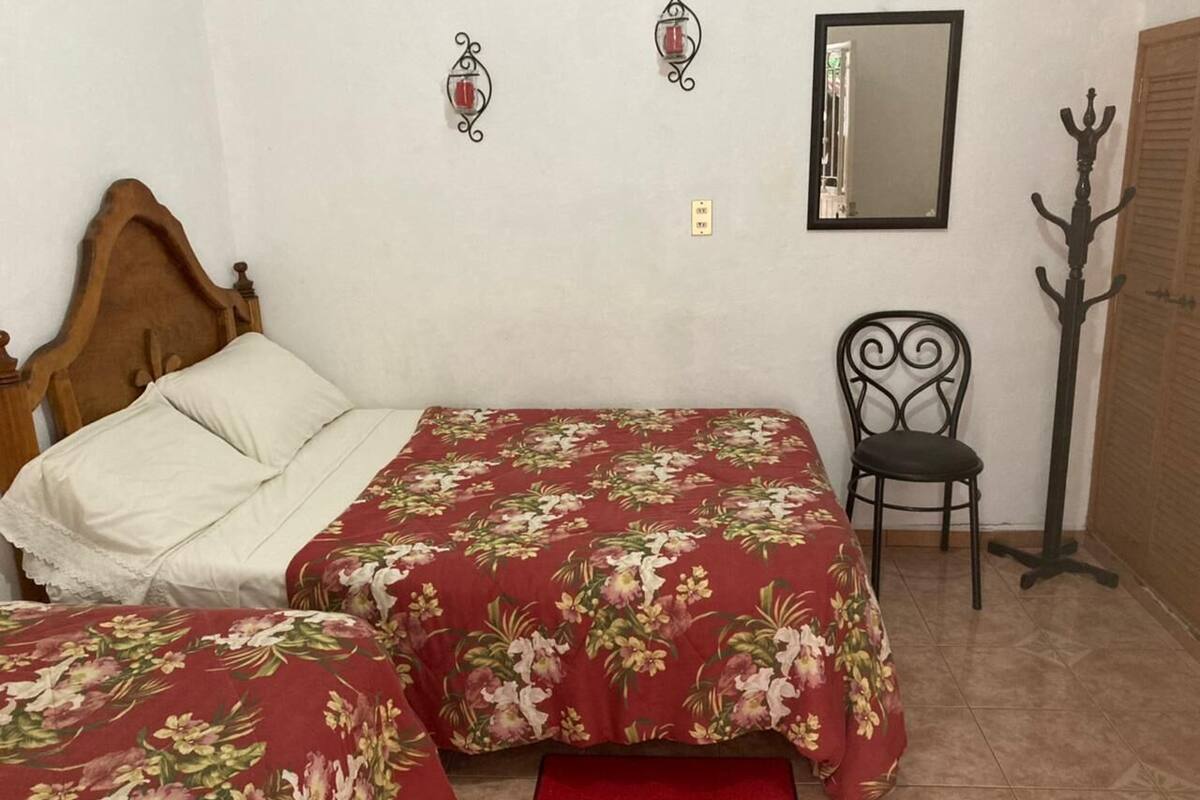 Quarto com cabeceira de madeira, duas camas com roupa de cama florida, uma cadeira, um porta-roupas e um espelho na parede.