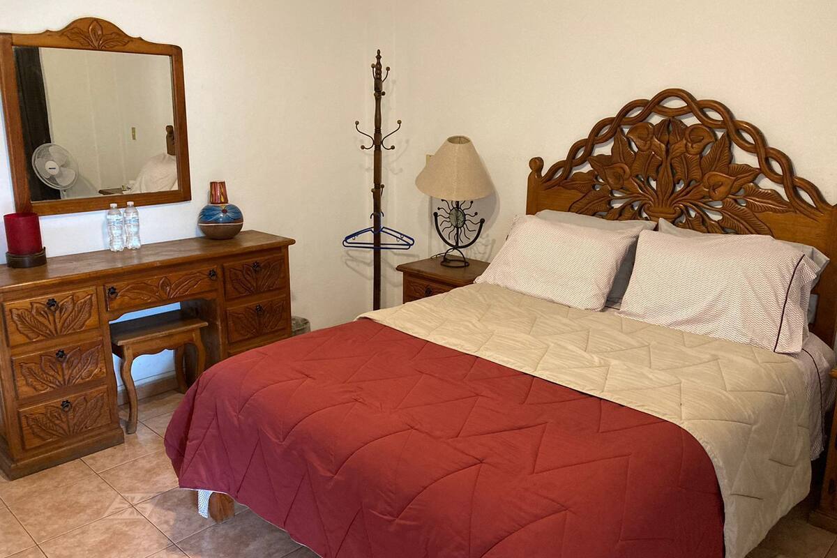 Quarto com cama de madeira, cómoda com gavetas, espelho, candeeiro e cabideiro.