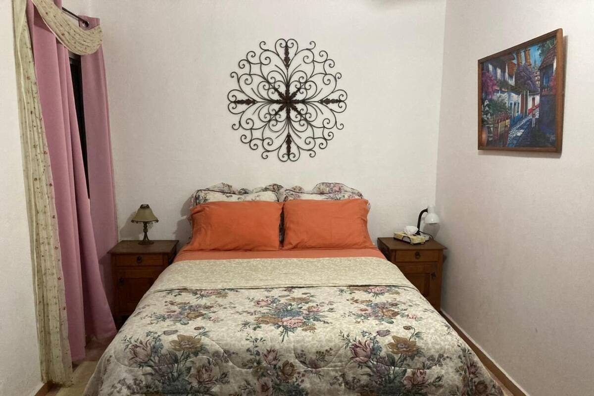 Um quarto com colcha florida, dois travesseiros laranja, um objeto decorativo na parede, uma mesinha de cabeceira com abajur e um quadro na parede.