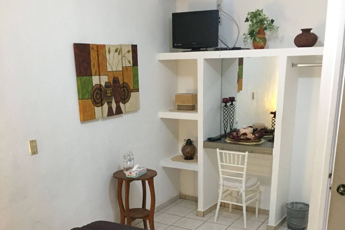 Quarto com cama, escrivaninha com cadeira, televisão e prateleira com objetos decorativos.