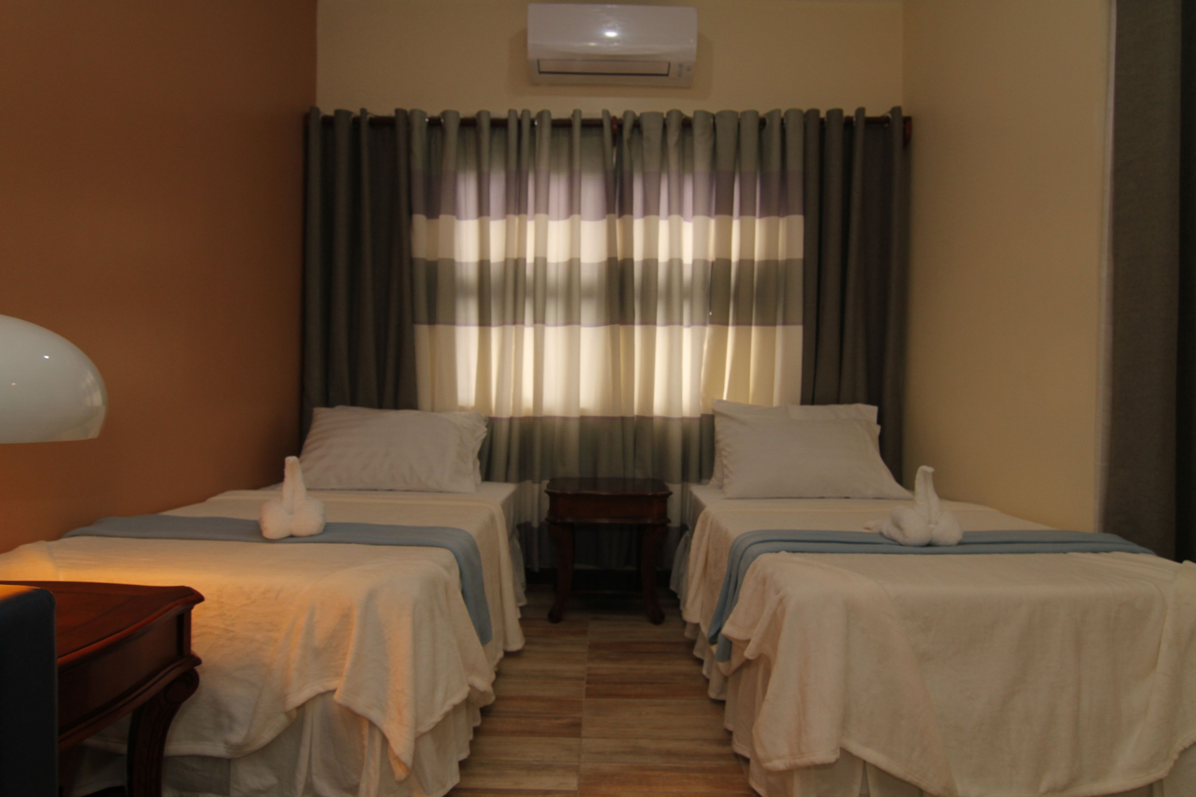 Quarto de hotel com duas camas, uma mesinha de cabeceira, um abajur e uma janela com cortinas.