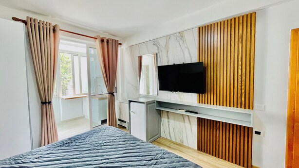 Quarto com cama, televisão fixada em painel de parede de madeira, pequena escrivaninha e janela com cortinas.