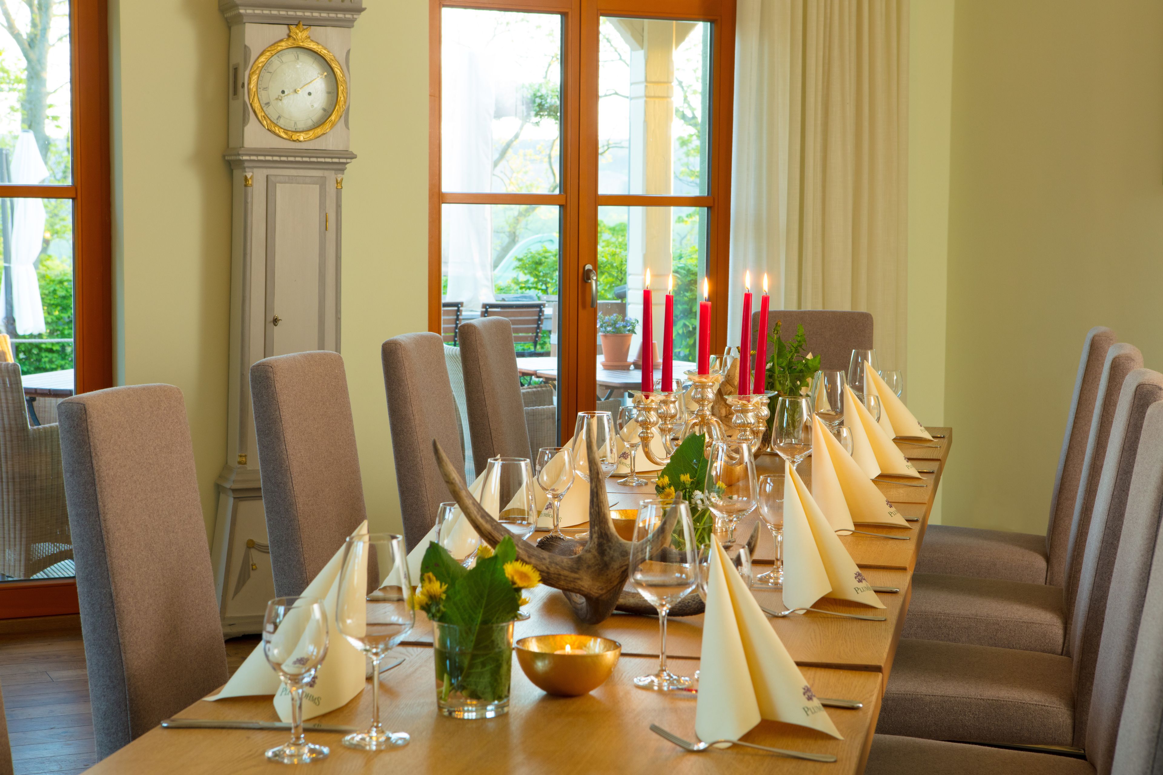 Mesa de jantar posta para uma refeição, com várias cadeiras, um relógio na parede e vista para o verde lá fora.