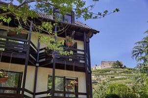 Um edifício com várias varandas, algumas com vasos suspensos. Vê-se uma colina com uma estrutura semelhante a um castelo ao fundo.