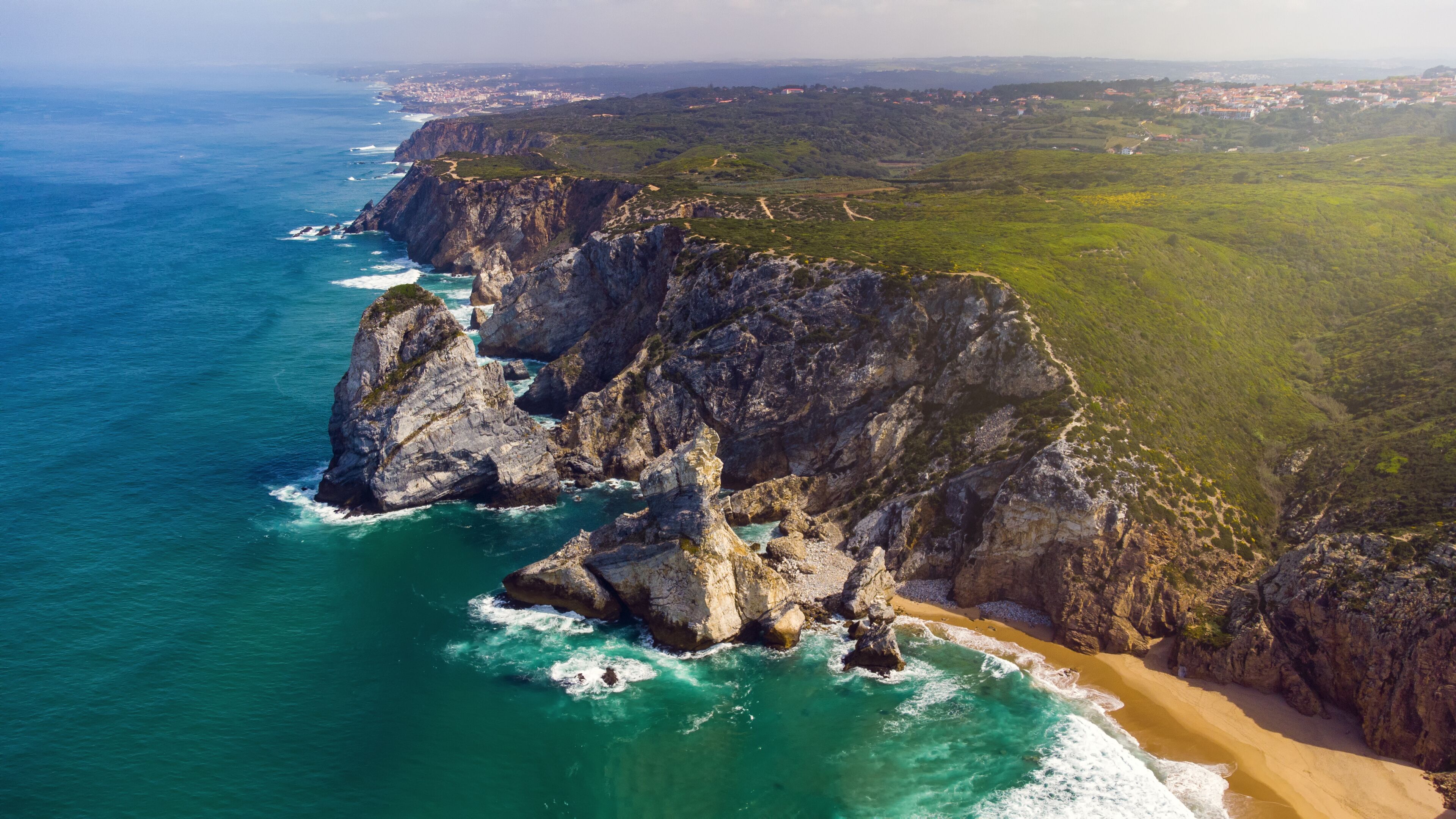 Paisagem costeira com falésias, uma praia e vista para uma cidade distante.