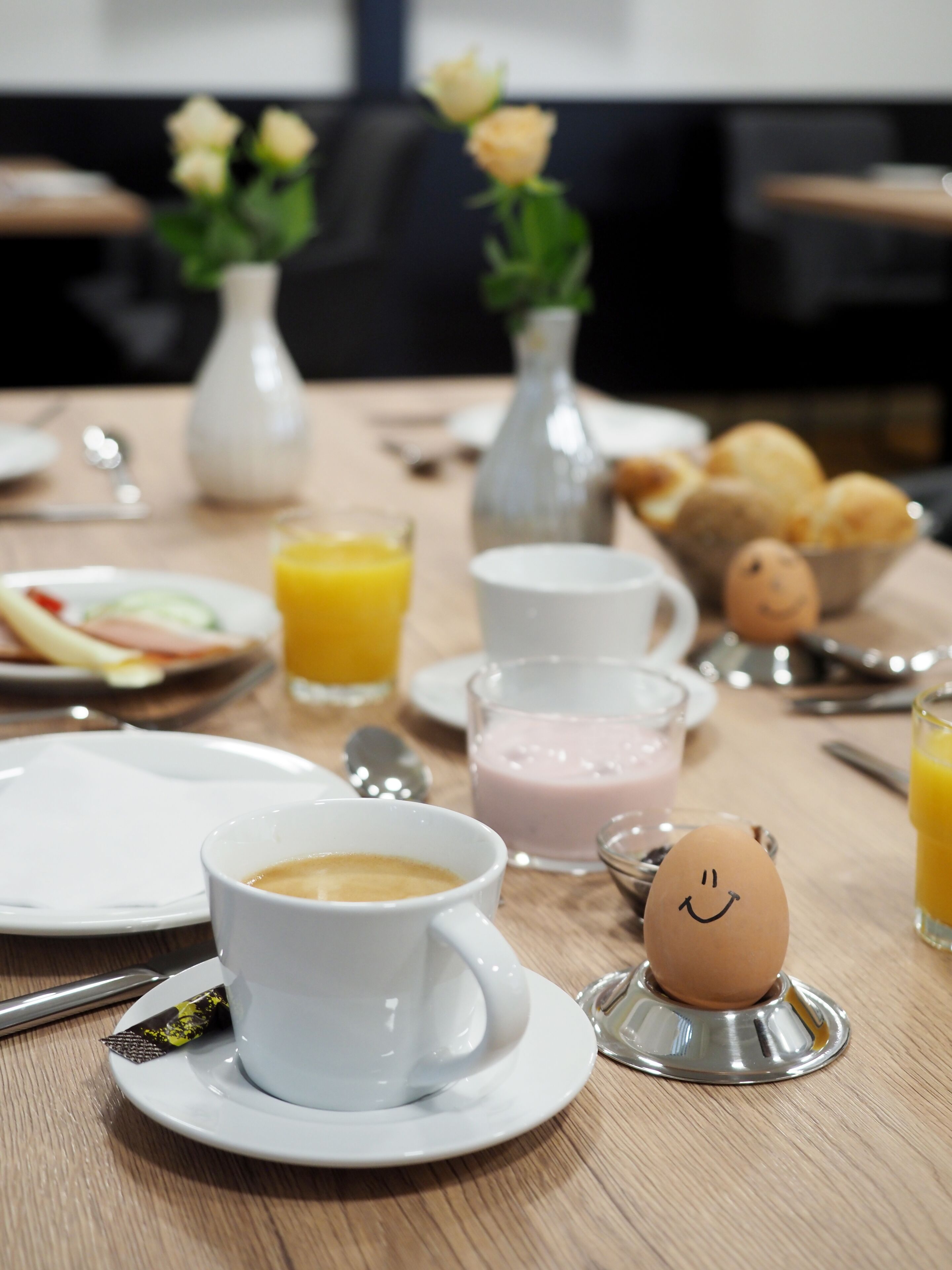 Uma mesa de café da manhã com uma xícara de café, um copo de suco de laranja, uma cesta de croissants e um ovo pequeno com carinha sorridente.