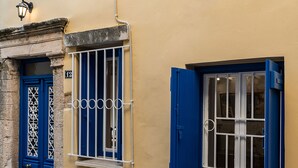Edifício com fachada amarela, portas azuis e janelas com barras brancas. Há plantas em vasos no chão.