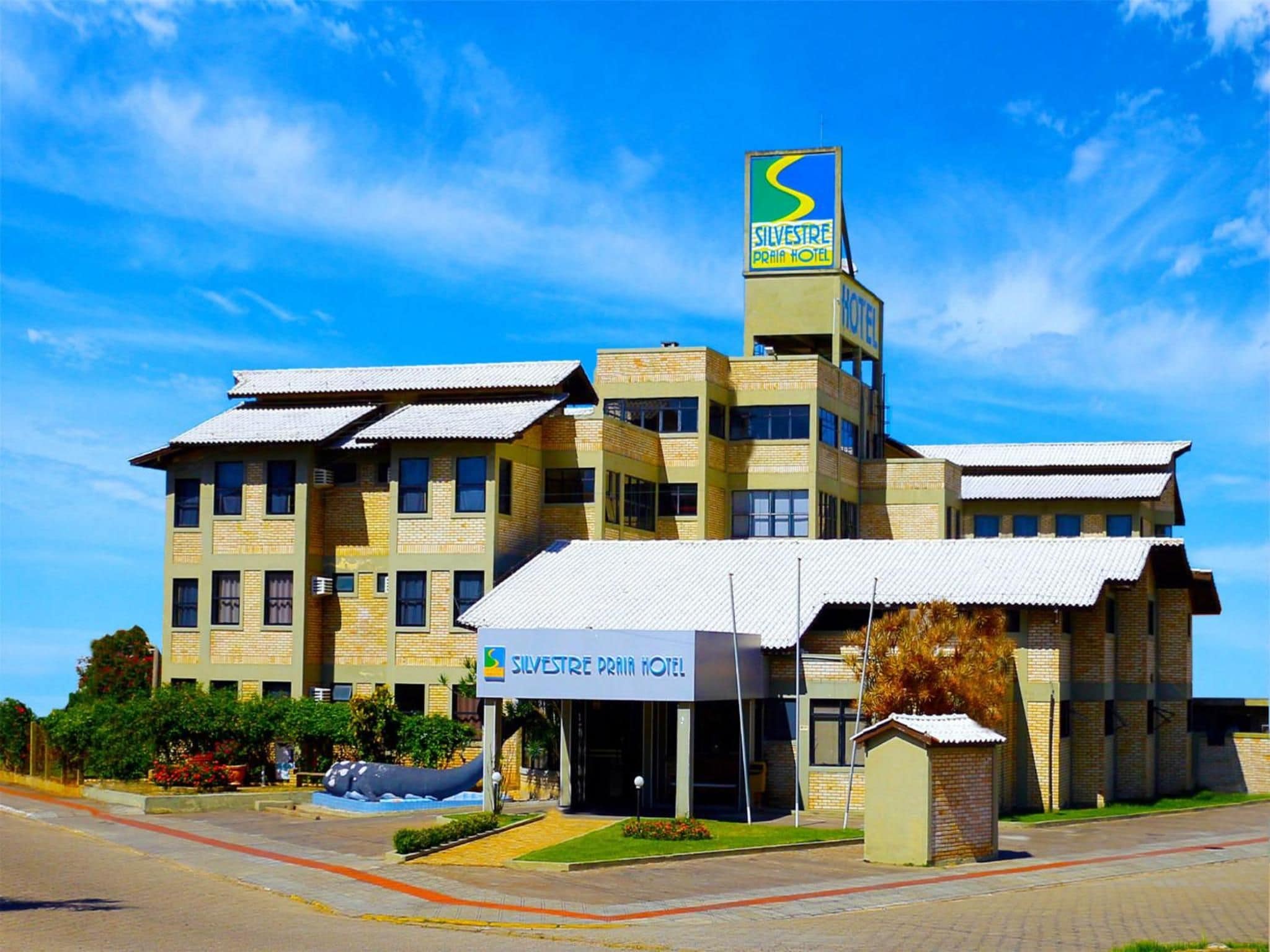 Um hotel com um letreiro grande de 'Silvestre Praia Hotel', vários andares e um estacionamento.