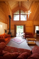 Interior de uma cabana de madeira com lareira, dois sofás, uma televisão e uma janela grande.