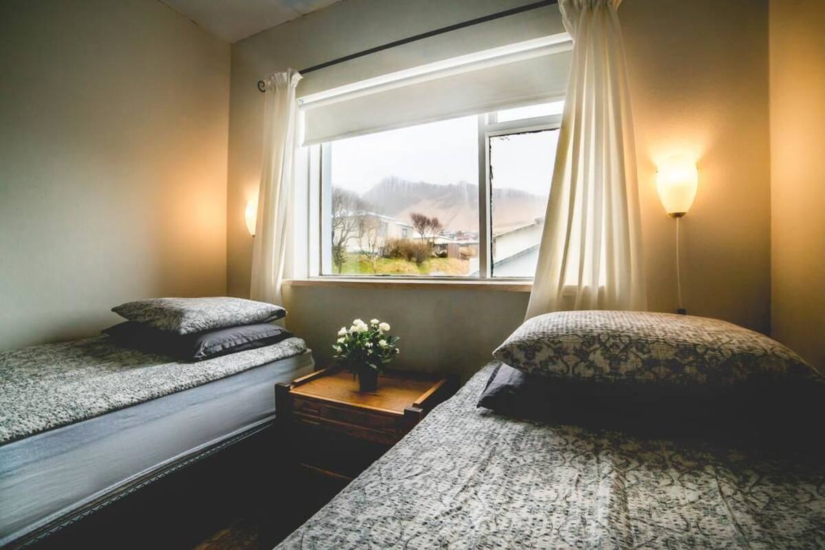 Quarto com duas camas de solteiro, janela com cortinas, mesa de cabeceira com um vaso de flores e uma luminária na parede.