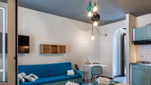 Quarto de hotel moderno com sofá azul, cama com colcha estampada, pequena mesa de jantar e cozinha compacta.