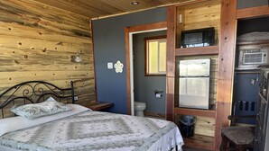 Um quarto com cama, mesa de cabeceira, cadeira, parede de madeira e banheiro visível através de uma porta aberta.