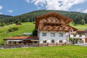 Um chalé de dois andares com varanda, rodeado por uma paisagem verdejante e uma montanha arborizada ao fundo.