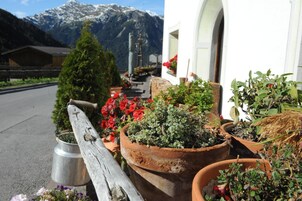 Um banco de madeira com vasos de plantas e flores, uma panela de metal e um edifício com fachada branca e entrada em arco.