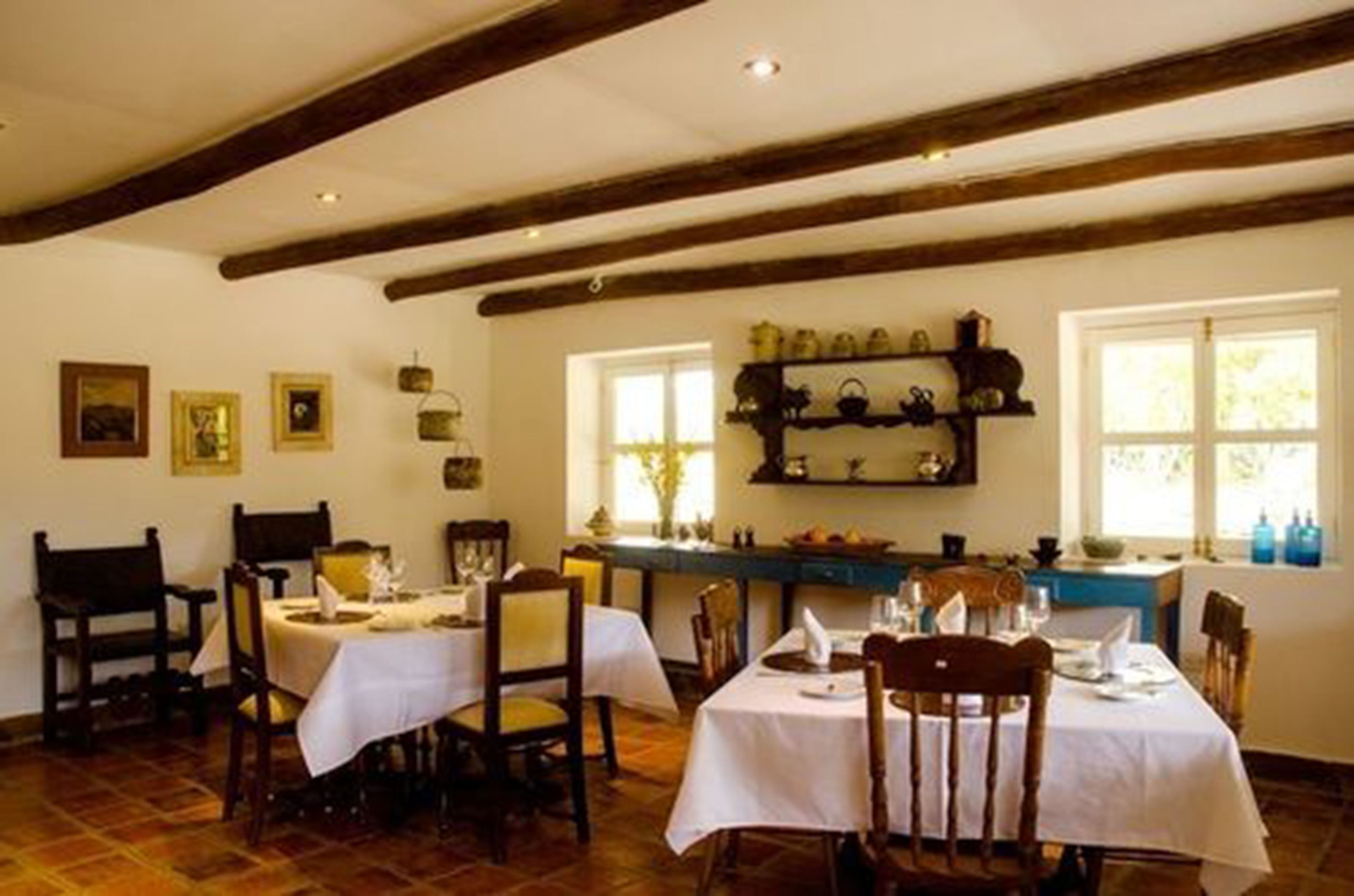 Uma área de refeições com vigas de madeira, mesas com toalhas brancas e uma estante com utensílios de cozinha.