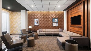 Um saguão de hotel com um sofá seccional, poltronas, uma televisão, uma parede estampada e um relógio.