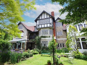Uma casa tradicional em estilo Tudor, com fachada de pedra, várias janelas e um jardim bem cuidado, com um banco e plantas em vasos.