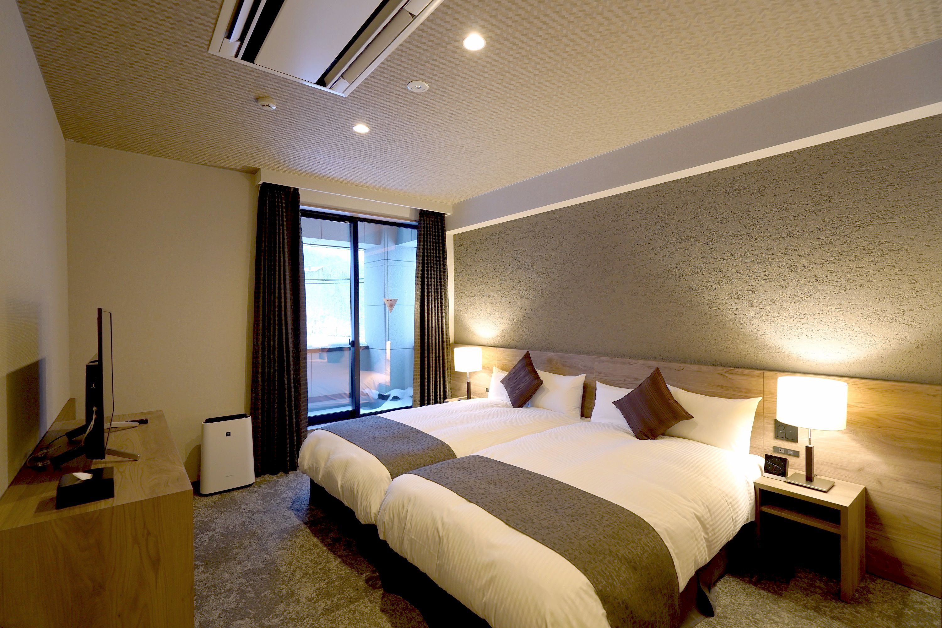 Quarto de hotel com duas camas, uma escrivaninha com computador, uma luminária e vista para o exterior.