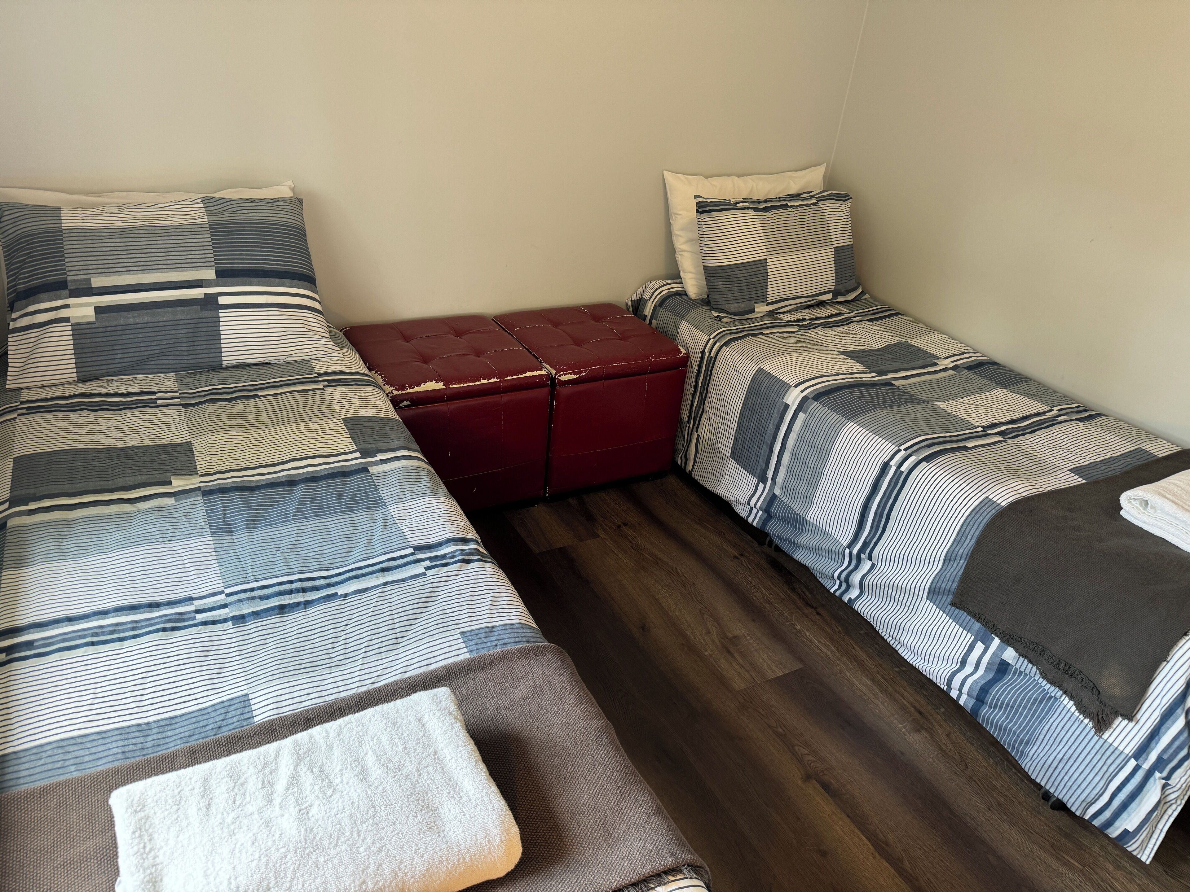 Quarto com duas camas, um banquinho vermelho e uma toalha branca no chão.