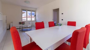 Uma sala de jantar moderna com uma mesa branca e cadeiras vermelhas, um sofá branco e uma janela com vista para edifícios.