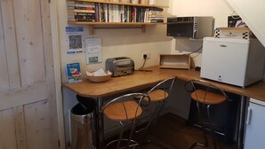 Uma cozinha compacta com bancada de madeira, dois bancos altos, uma torradeira, um micro-ondas e uma estante cheia de livros.