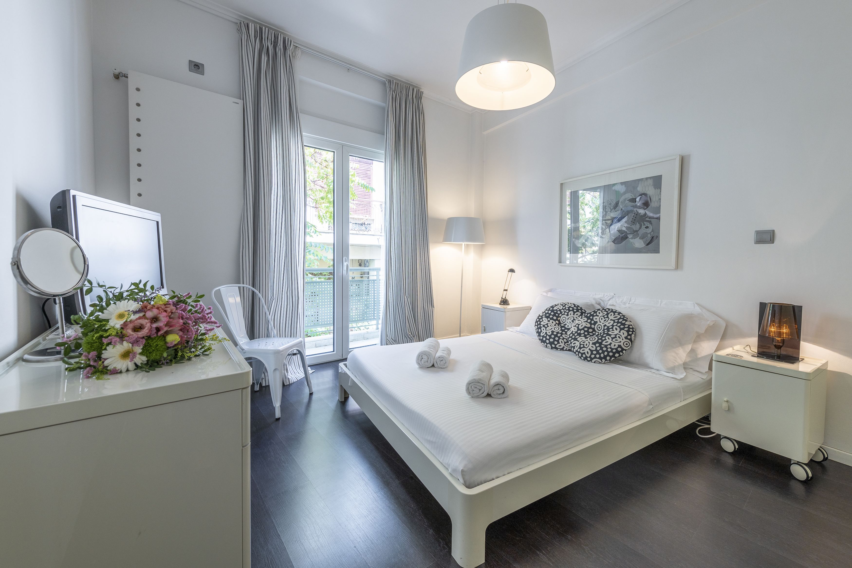 Quarto moderno com cama, mesa de cabeceira, abajur e escrivaninha com espelho e flores.