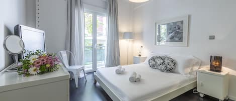 Quarto moderno com cama, mesa de cabeceira, abajur e escrivaninha com espelho e flores.