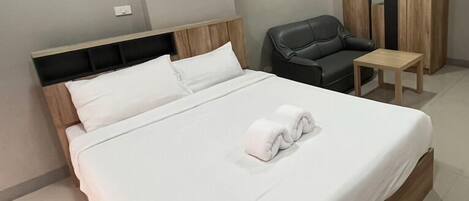 Cama bem arrumada com lençóis brancos e uma toalha enrolada, um pequeno criado-mudo de madeira, uma poltrona de couro e um guarda-roupa com listras verticais.