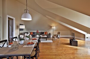 Um loft espaçoso com pé-direito alto, piso de madeira, área de jantar com uma mesa comprida e um espaço aconchegante para relaxar com sofá e TV.