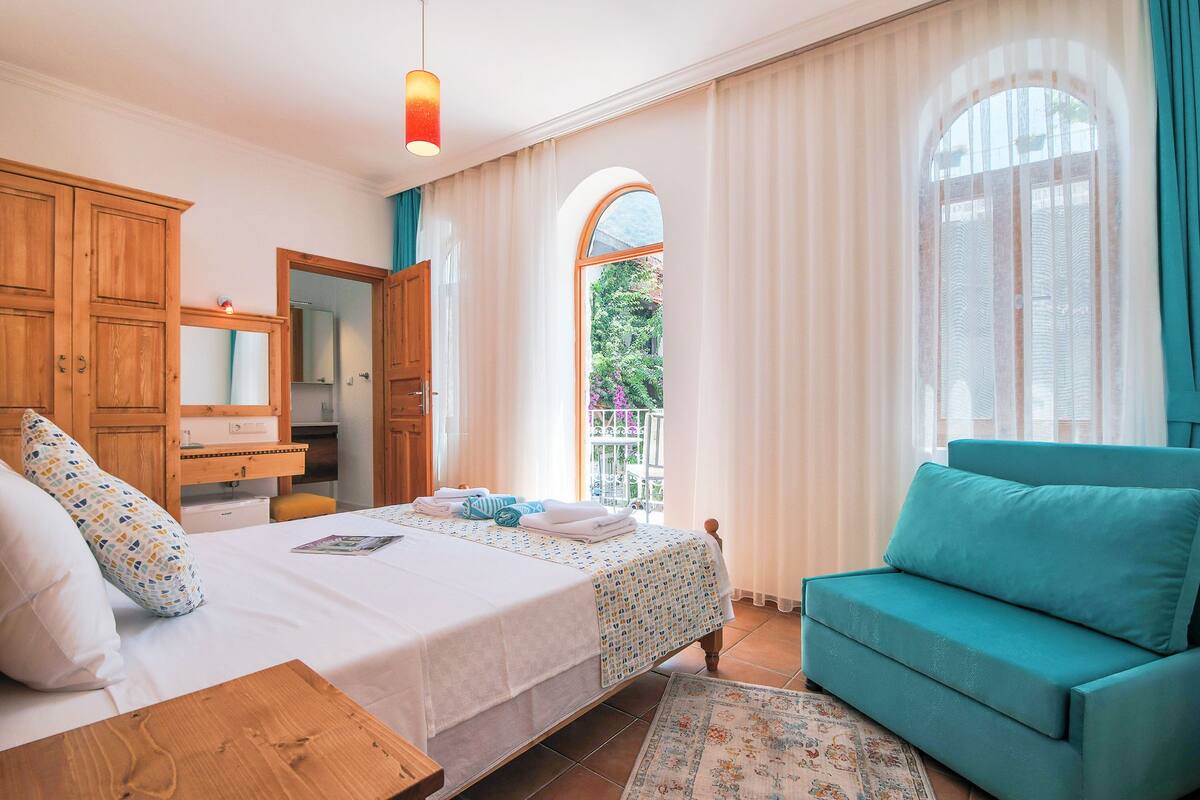 Quarto com cama, guarda-roupa de madeira, sofá verde-água e janela com cortinas.