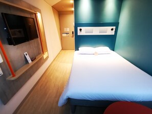 Quarto de hotel com cama, televisão fixada na parede, piso de madeira e parede azul decorativa.