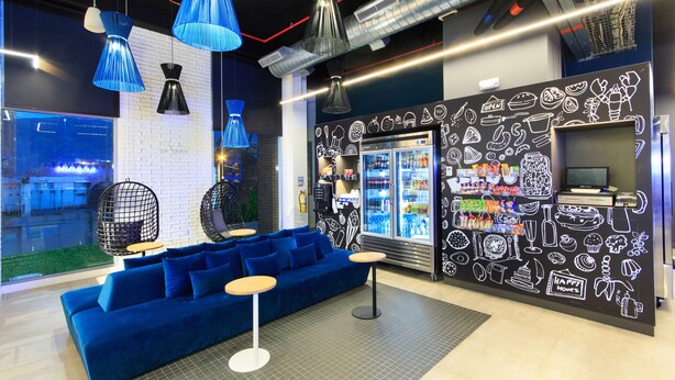 Uma área de estar moderna com um sofá azul, luminárias pendentes, uma geladeira repleta de lanches e uma parede de lousa com ilustrações de comidas.