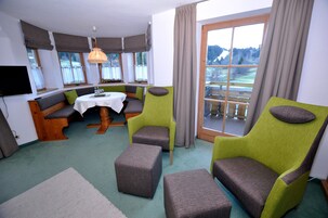 Um quarto aconchegante com uma mesa de jantar, duas poltronas verdes, um banco e vista para uma paisagem nevada por uma grande janela.