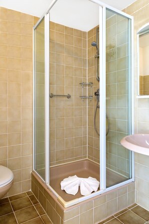 Casa de banho com chuveiro em cabine de vidro, azulejos bege, lavatório branco e uma toalha no chão.
