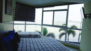 Quarto com uma grande janela com vista para um corpo de água, uma cama com colcha xadrez e uma televisão presa à parede.