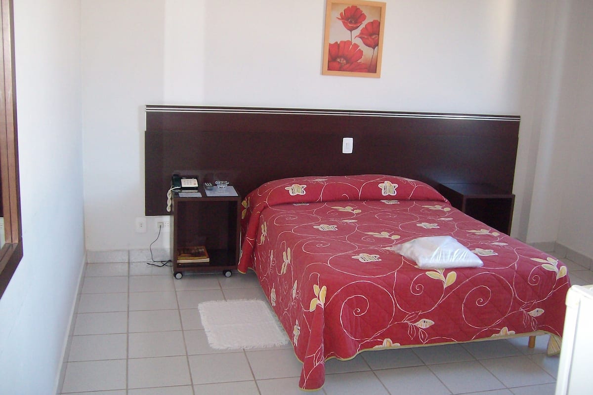 Quarto com uma cama, mesa de cabeceira, telefone fixado na parede e um quadro enquadrado.