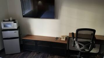 Quarto de hotel com cama, escrivaninha com cadeira, micro-ondas, frigorífico e televisão fixada na parede.