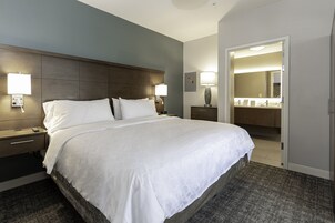 Quarto de hotel com uma cama grande, luminárias de cabeceira, mesa de cabeceira, banheiro com espelho e porta que dá para outro quarto.