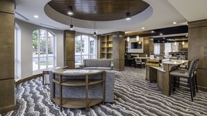 Um hall de hotel moderno com teto circular de madeira, grandes janelas, área de estar com mesa redonda e cozinha ao fundo.