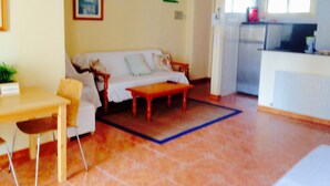 Sala de estar com sofá, mesa de centro de madeira e área de refeições com mesa e cadeiras. É visível uma cozinha ao fundo.