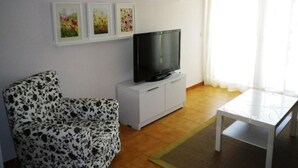 Uma sala de estar com sofá, televisão em um móvel, uma mesa e quadros na parede.