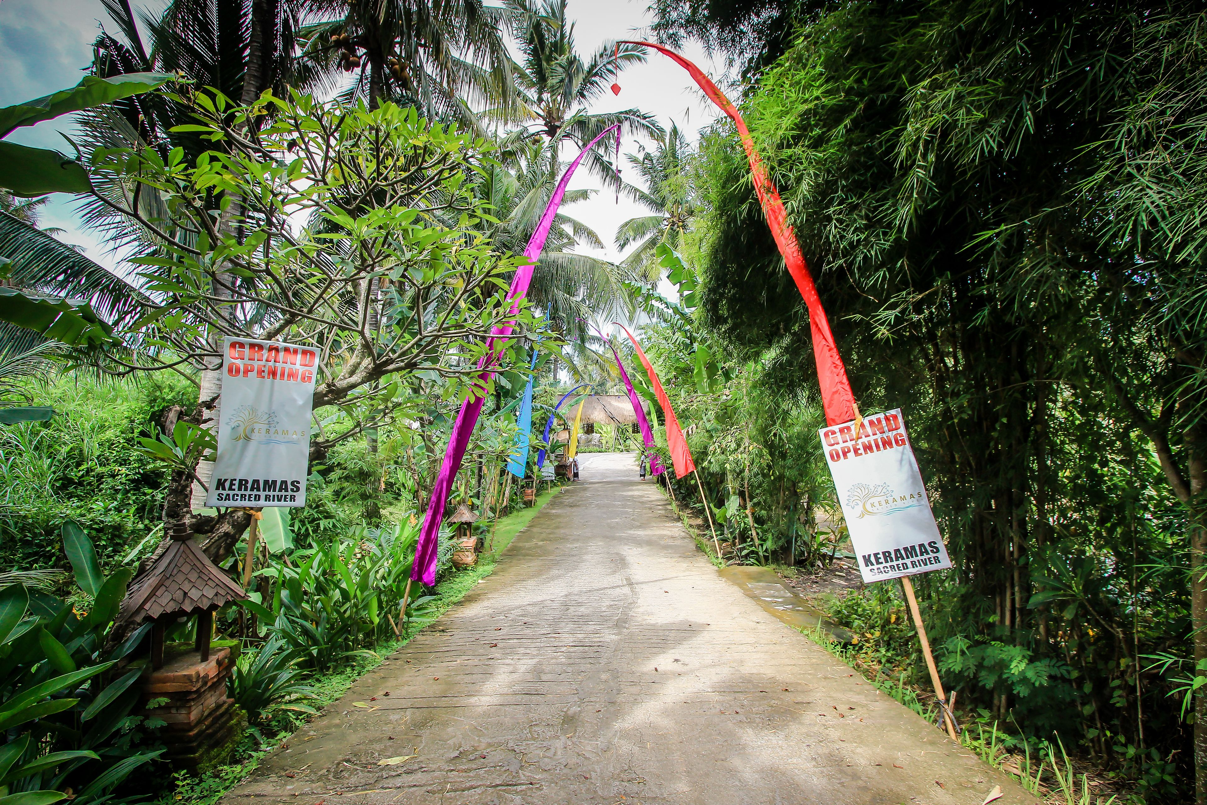 Uma trilha decorada com bandeiras coloridas e placas indicando a inauguração de 'Keramas Sacred River'.