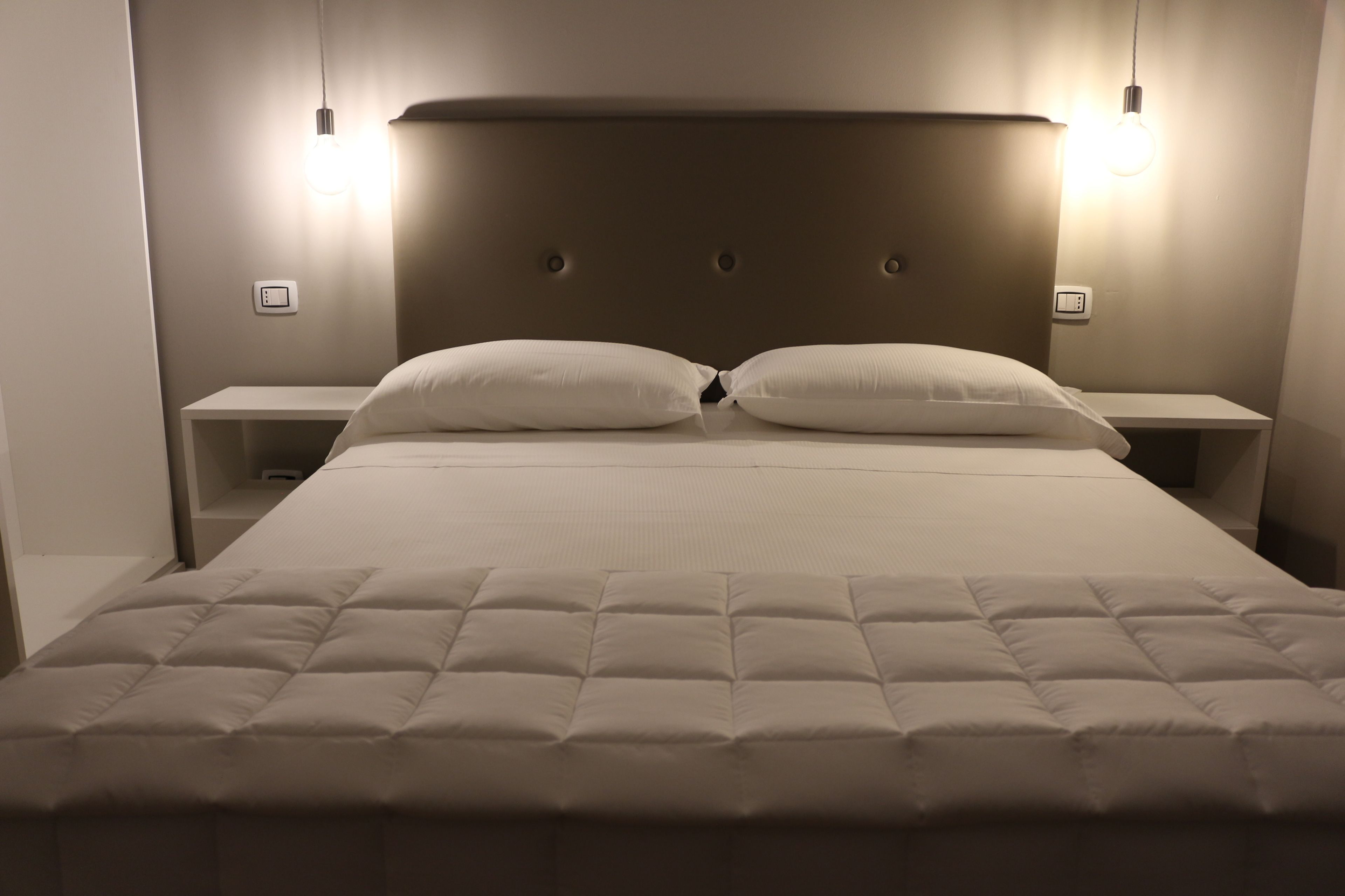 Uma cama bem arrumada com cabeceira estofada, duas mesas de cabeceira e duas luminárias fixadas na parede.