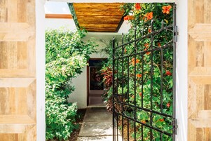 Uma entrada estreita com portão de metal, cercada por vegetação exuberante e plantas floridas.