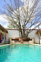 Piscina com uma árvore ao fundo, uma rede e um edifício com telhado de palha.
