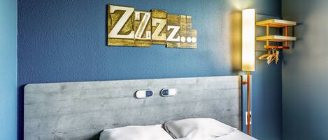 Quarto de hotel com parede azul, cabeceira cinzenta, cama com roupa de cama branca, luminária de parede, e prateleira com abajur e chaveiro.