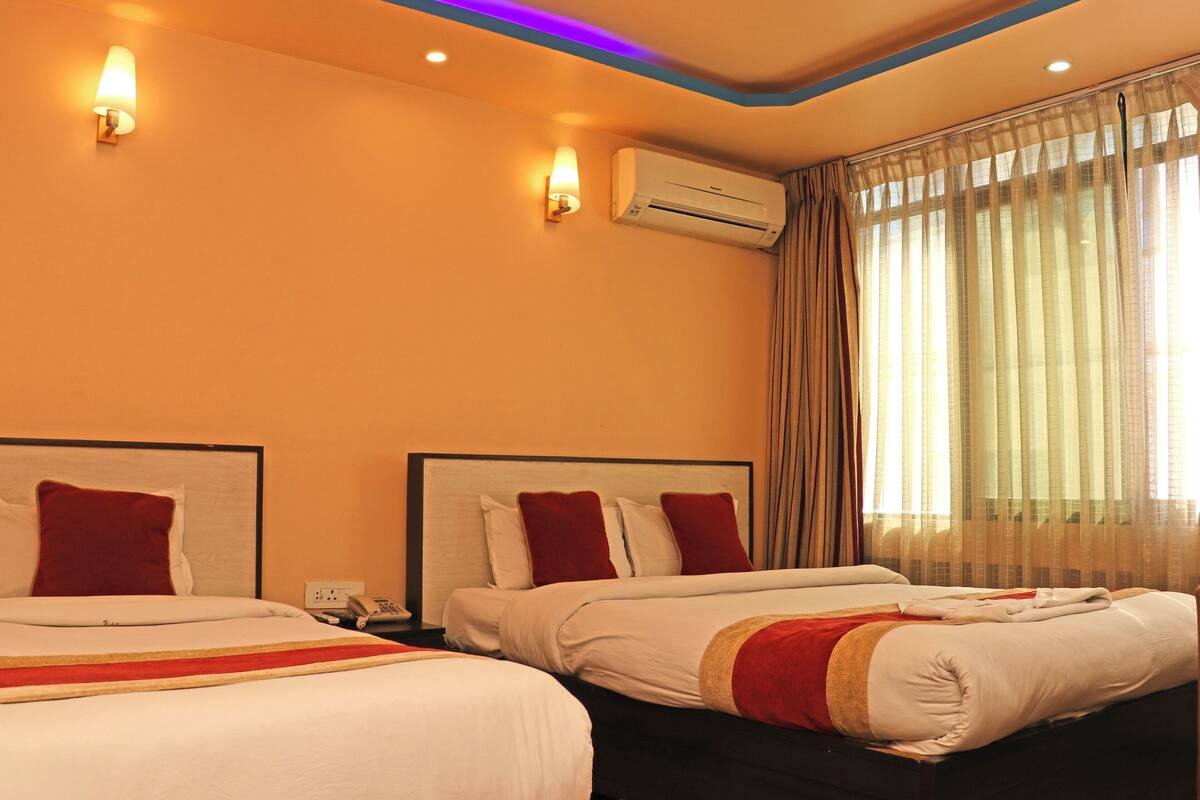 Quarto de hotel com duas camas, ar-condicionado fixado na parede e uma janela com cortinas leves.
