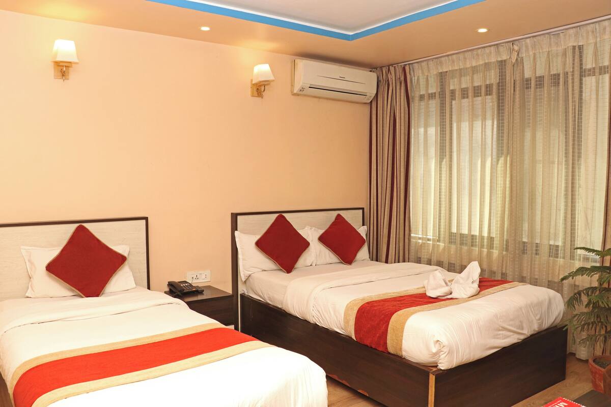 Quarto de hotel com duas camas, cada uma com almofadas vermelhas, um cobertor listrado em vermelho e branco e uma toalha branca sobre a cama. Há um ar condicionado na parede e uma planta no canto.